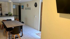 Uma sala de jantar com uma mesa e cadeiras de madeira, um ventilador de teto e uma cozinha ao fundo.