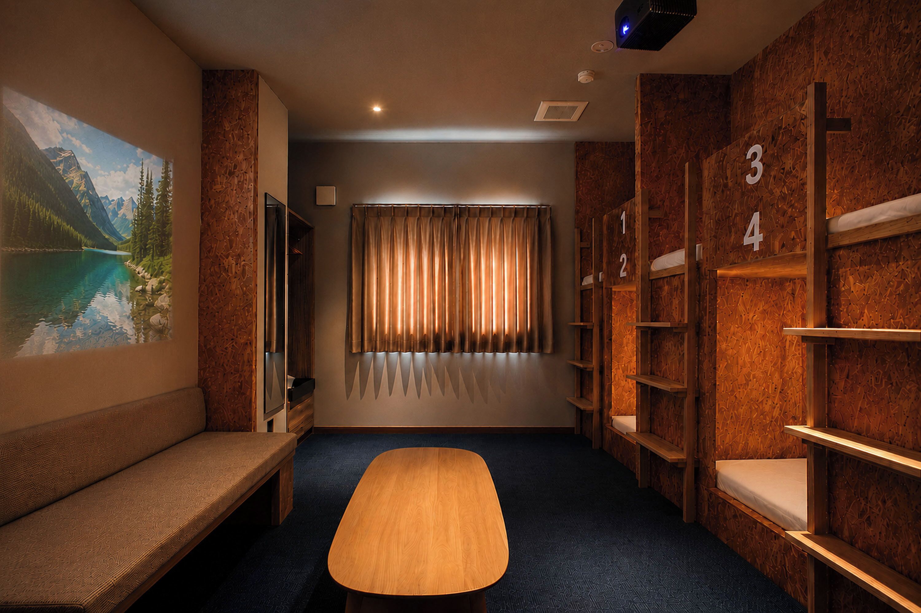 Quarto de hotel com uma pintura grande, um banco de madeira, beliches e uma tela de projeção.