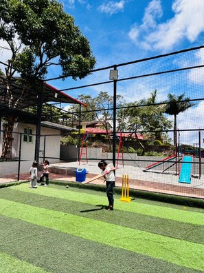 Uma área de treino de críquete ao ar livre, com uma rede delimitadora, uma jaula de rebatedores e um pequeno pavilhão.
