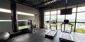 Uma academia com equipamentos de exercício, um saco de pancada e vista para o horizonte urbano através de grandes janelas.
