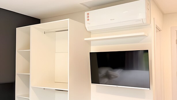 Quarto com um guarda-roupa branco, ar condicionado fixo e uma televisão sobre um suporte.