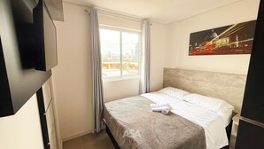 Quarto de hotel com cama, televisão, janela com cortinas e vista para a cidade.