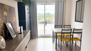 Uma área de refeições com mesa e cadeiras, uma janela grande com cortinas, e uma parede com um quadro e um relógio.