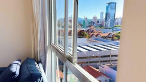 Quarto com uma grande janela, uma cama com almofadas e vista para um horizonte urbano com edifícios.