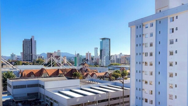 Um edifício alto com várias varandas, céu claro e paisagem urbana com diversos edifícios ao fundo.