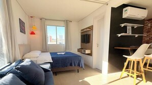 Quarto de hotel moderno com cama, sofá, escrivaninha com duas cadeiras, televisão e parede de tijolos.