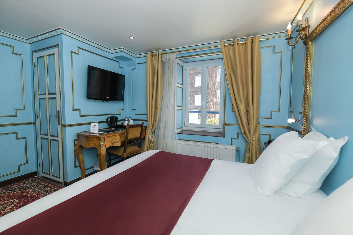 Quarto de hotel com cama, escrivaninha, televisão e janela com cortinas.
