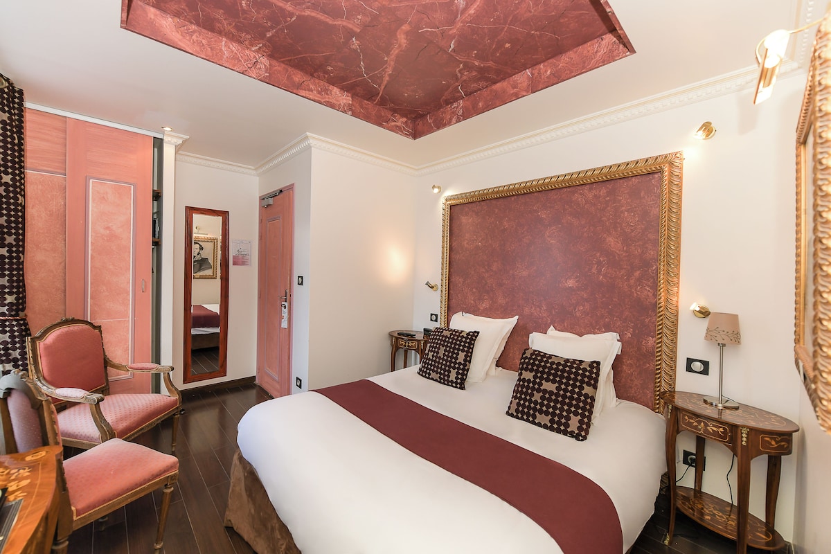 Quarto de hotel com uma cama grande, área de estar com uma poltrona, espelho e cabeceira de madeira.
