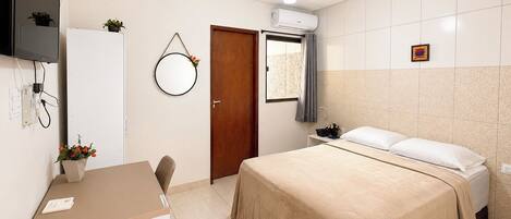 Quarto de hotel com cama, mesa de cabeceira, cadeira, escrivaninha, televisão, aparelho de ar condicionado e janela com cortinas.