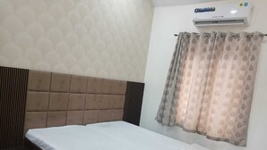 Uma cama de solteiro com cabeceira, ar condicionado fixado na parede e uma janela com cortinas estampadas.