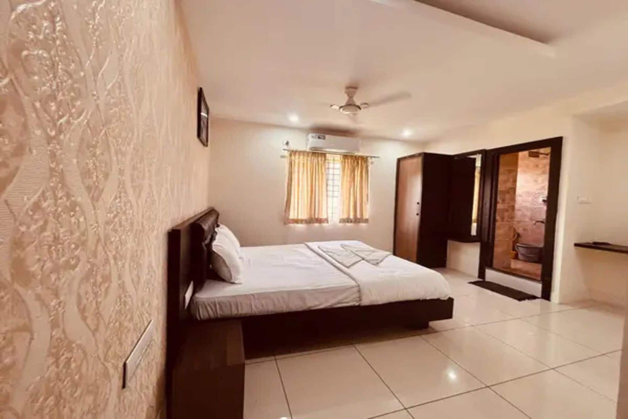 Quarto com cama, ventoinha no teto, janela com cortinas e banheiro visível por uma porta aberta.