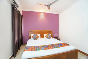 Quarto com cama de madeira, paredes roxas, ventilador de teto e janela com cortinas.