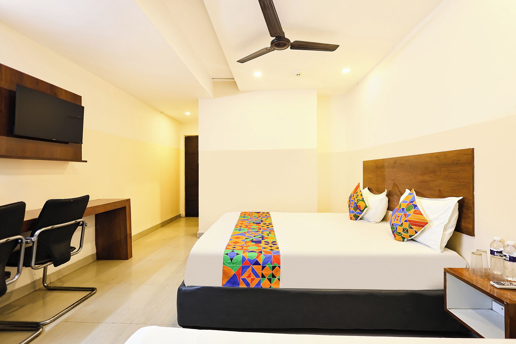 Quarto de hotel com cama, escrivaninha, cadeira, televisão e ventoinha no teto.