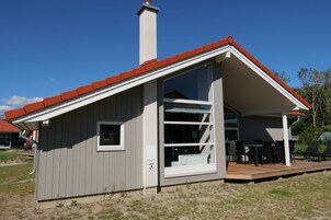 Um pequeno edifício térreo com telhado vermelho, uma grande porta de correr de vidro e uma pequena área de deck com móveis de exterior.