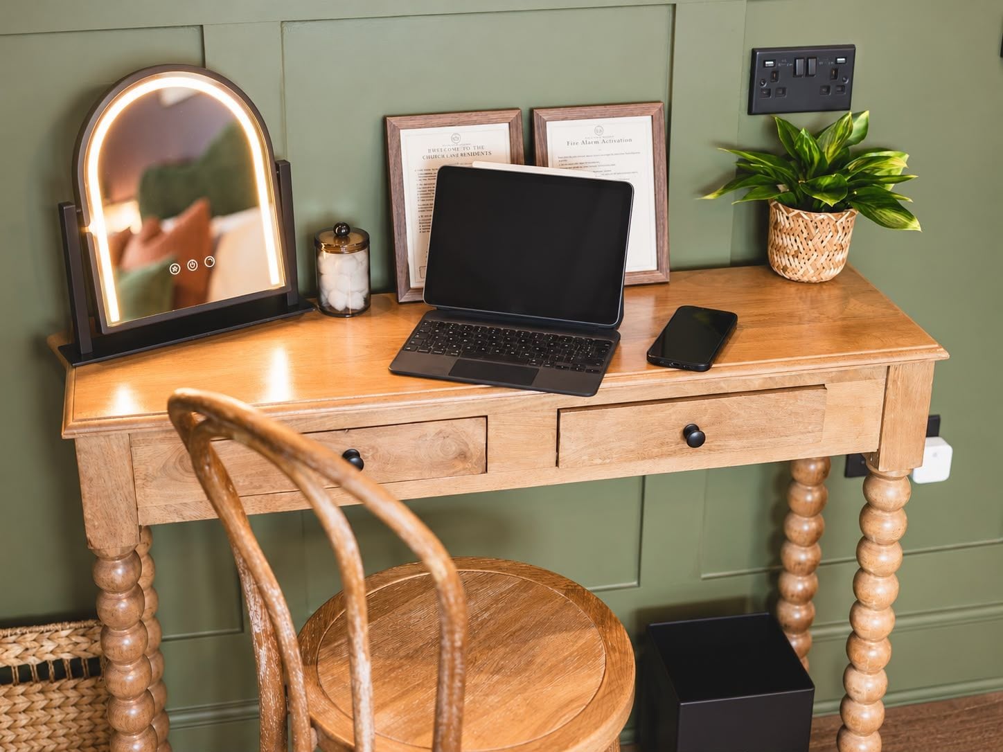 Mesa de madeira com um laptop, um smartphone, um certificado emoldurado, uma pequena planta em vaso e um espelho com luz.