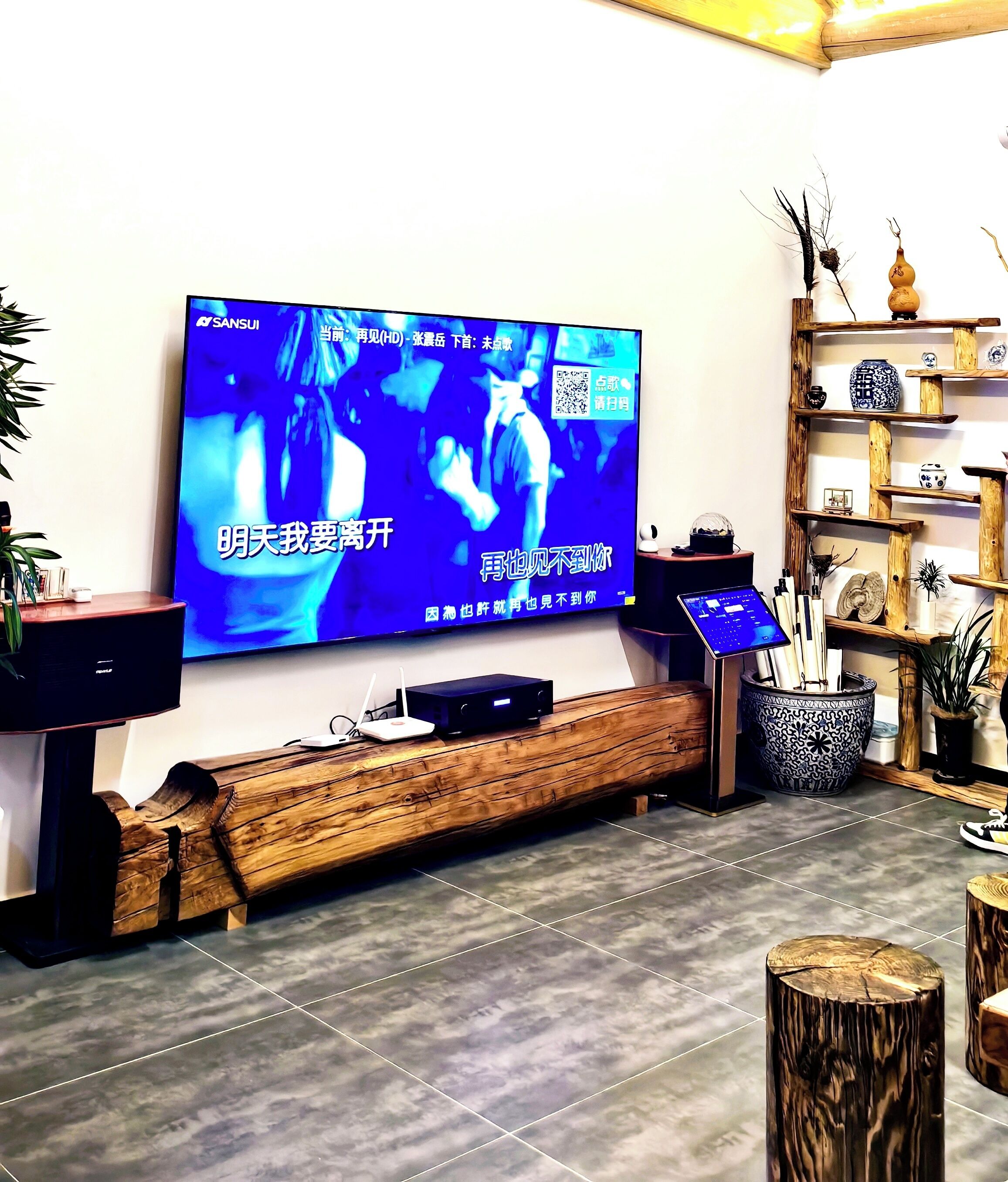 Uma sala de estar moderna com uma televisão de tela plana na parede, um banco de madeira e uma estante decorativa com diversos objetos.