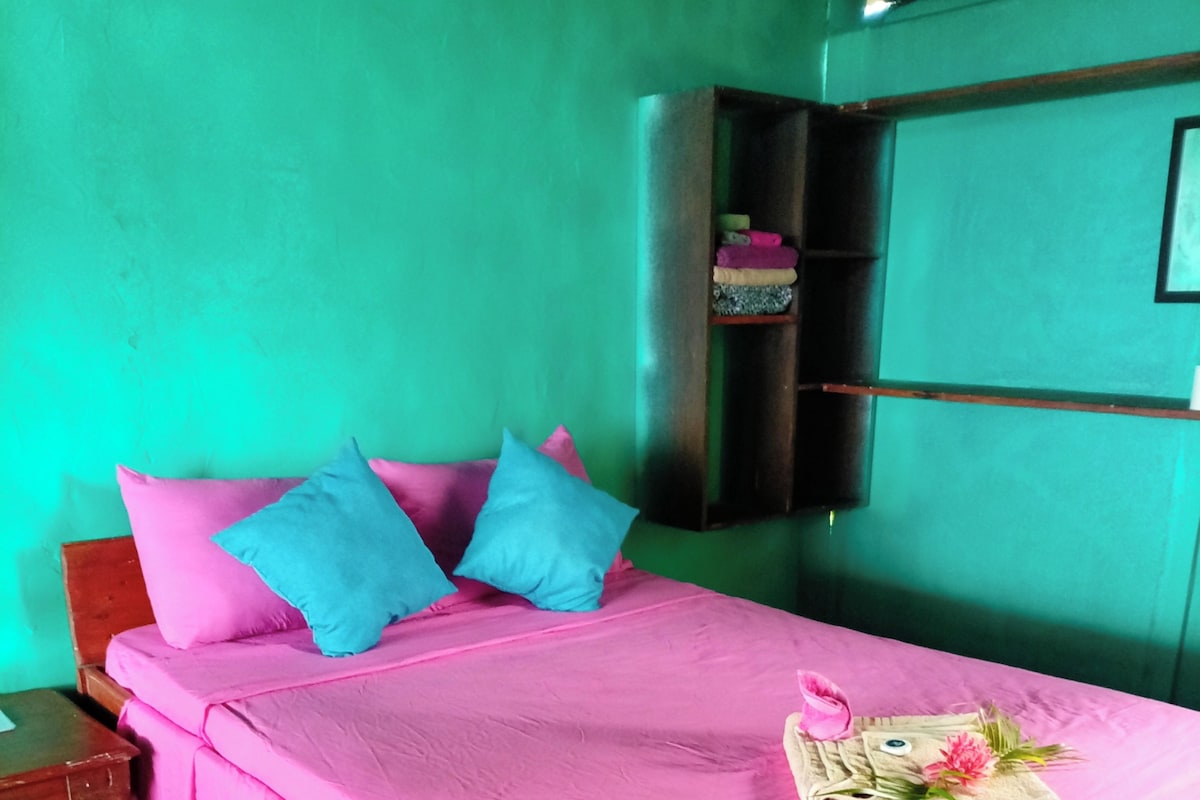 Cama com roupa de cama rosa e almofadas azuis, mesa de cabeceira de madeira e uma pequena estante com roupas dobradas.