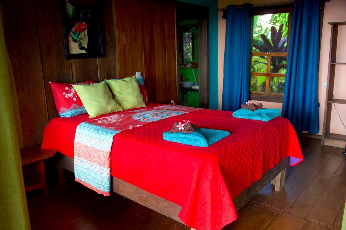 Uma cama com colcha e almofadas vermelhas, chão de madeira, janela com cortinas e uma pequena mesa com uma planta.