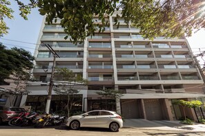 Um edifício com vários andares, varandas, área de estacionamento com carros e motocicletas, e entrada com portão.