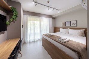 Quarto de hotel com cama, escrivaninha, televisão e janela com cortinas.