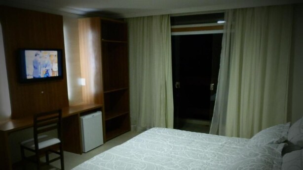 Quarto de hotel com cama, escrivaninha, cadeira e televisão.