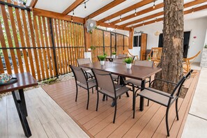 Um pátio externo com mesa e cadeiras de madeira, uma área de recepção e uma árvore.