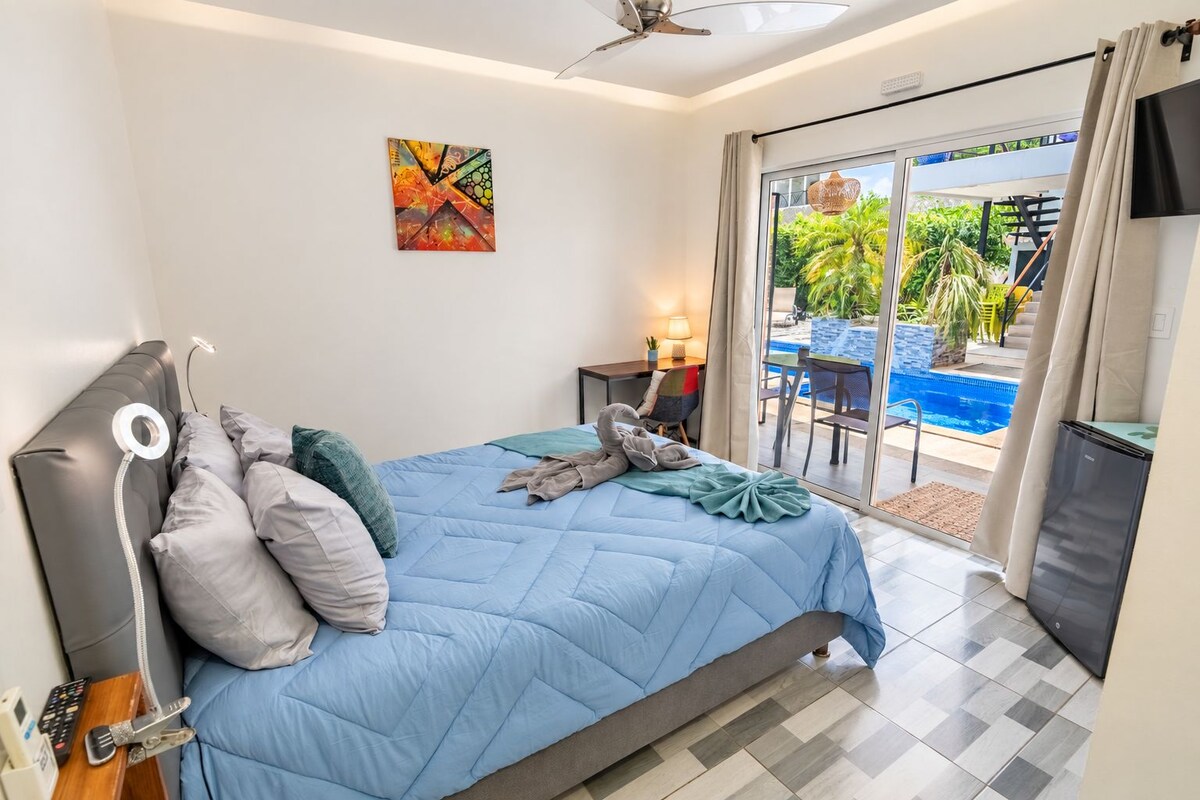 Quarto com cama, escrivaninha, cadeira, televisão e vista para uma piscina exterior.