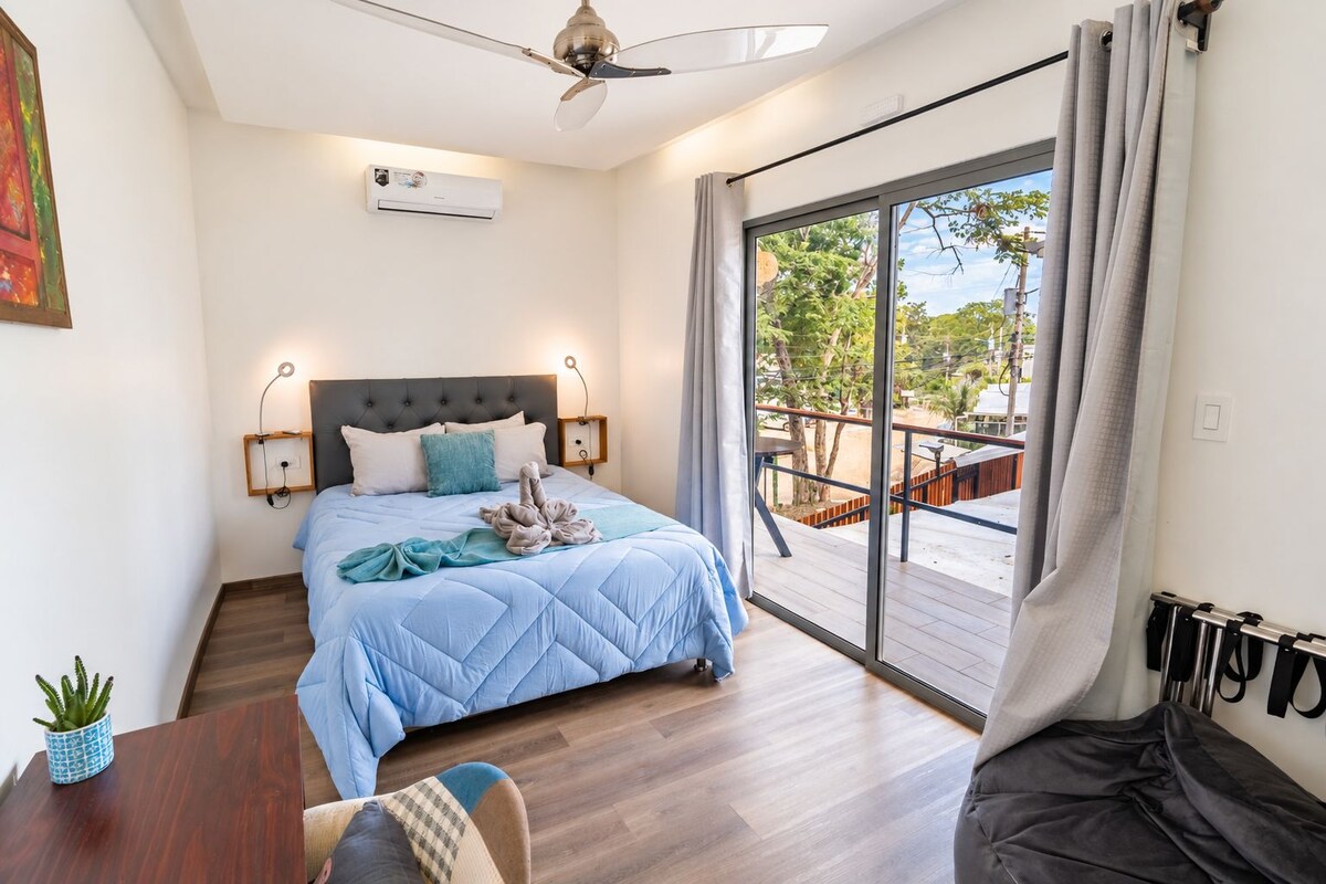 Quarto com cama, ventoinha no teto, janela com vista para o exterior e uma pequena mesa com uma planta.