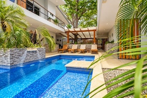 Uma piscina moderna ao ar livre com borda de pedra, rodeada por espreguiçadeiras e plantas em vasos.