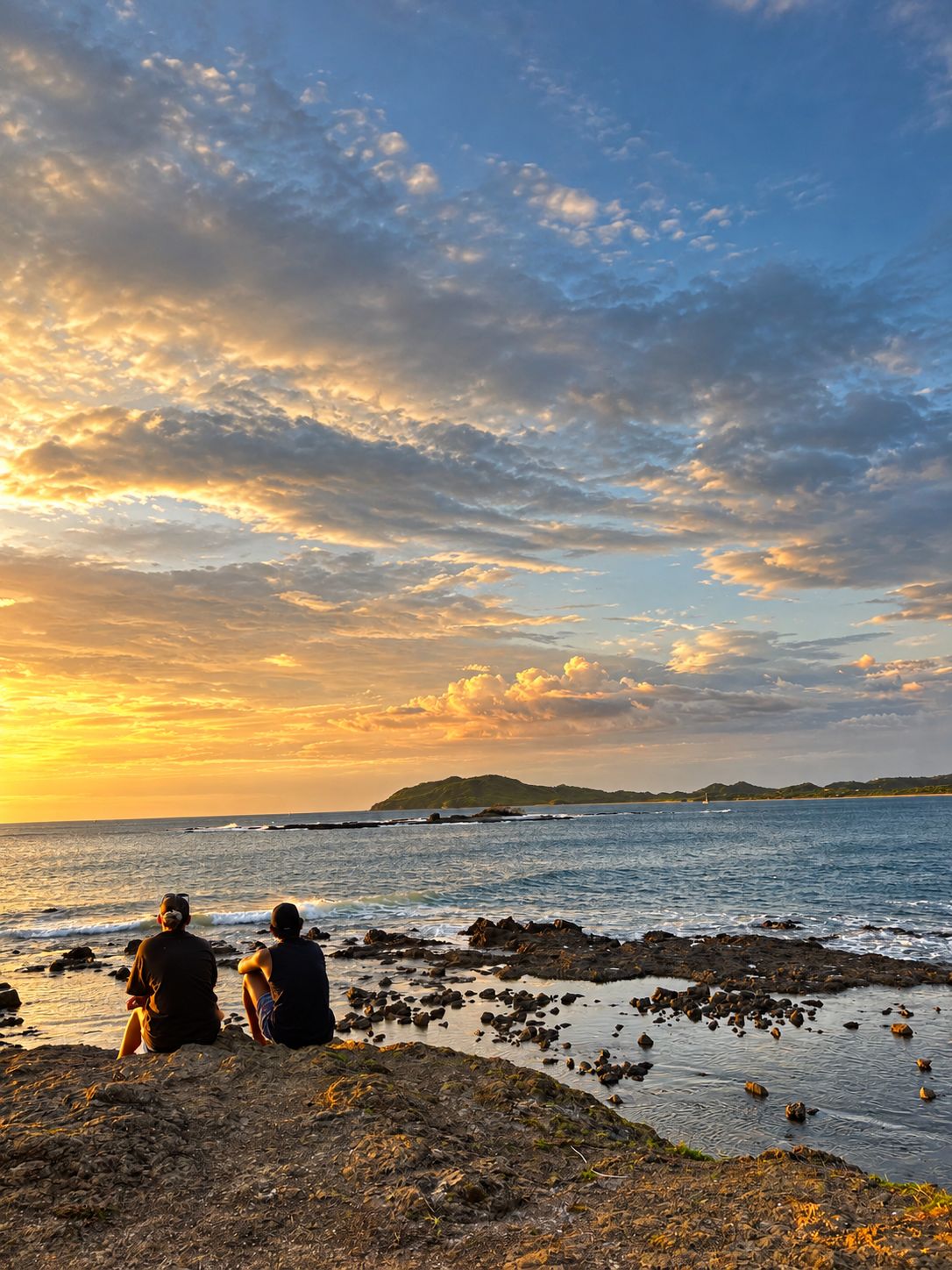 Duas pessoas sentadas em uma praia rochosa, com vista para o mar e a terra ao longe durante o pôr do sol.