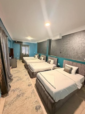 Quarto de hotel com quatro camas dispostas em linha, uma escrivaninha e uma janela grande com cortinas.