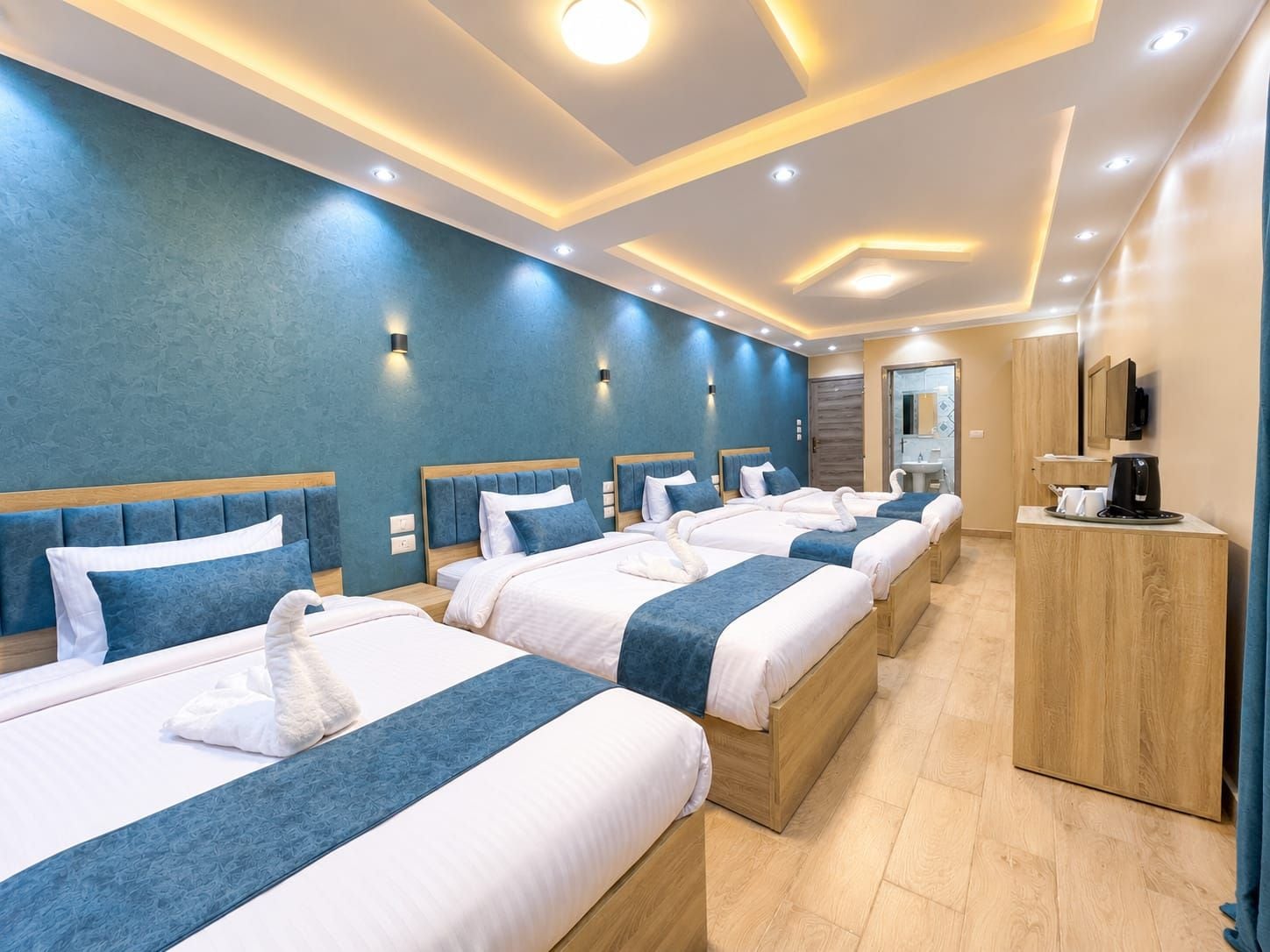 Quarto de hotel com quatro camas dispostas em linha, um armário de madeira e um espelho na parede.