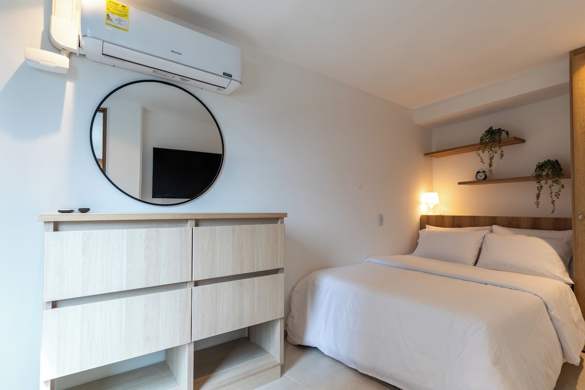 Quarto com cama, mesa de cabeceira, espelho, ar-condicionado na parede e prateleira com plantas.
