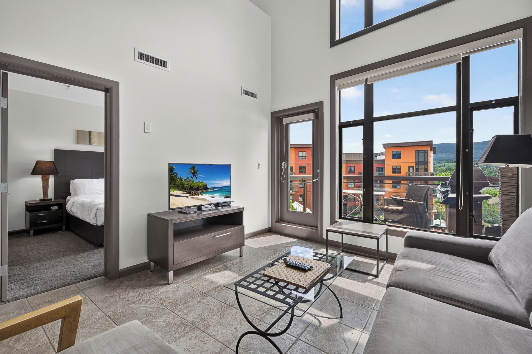 Quarto de hotel moderno com cama, televisão de tela plana, mesa de centro e janelas amplas com vista para edifícios e uma praia.