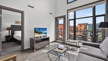 Quarto de hotel moderno com cama, televisão de tela plana, mesa de centro e janelas amplas com vista para edifícios e uma praia.