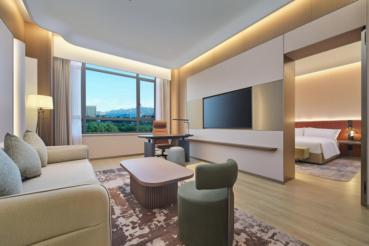 Quarto de hotel moderno com sofá, poltrona, mesa de centro e televisão fixada na parede.