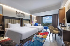 Quarto de hotel com duas camas, uma escrivaninha, uma cadeira, televisão e uma janela grande com vista para a cidade.