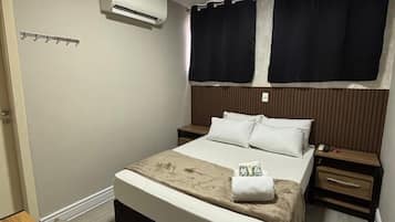 Quarto com cama, mesas de cabeceira, ar condicionado, janela com cortinas e uma pequena mesa.