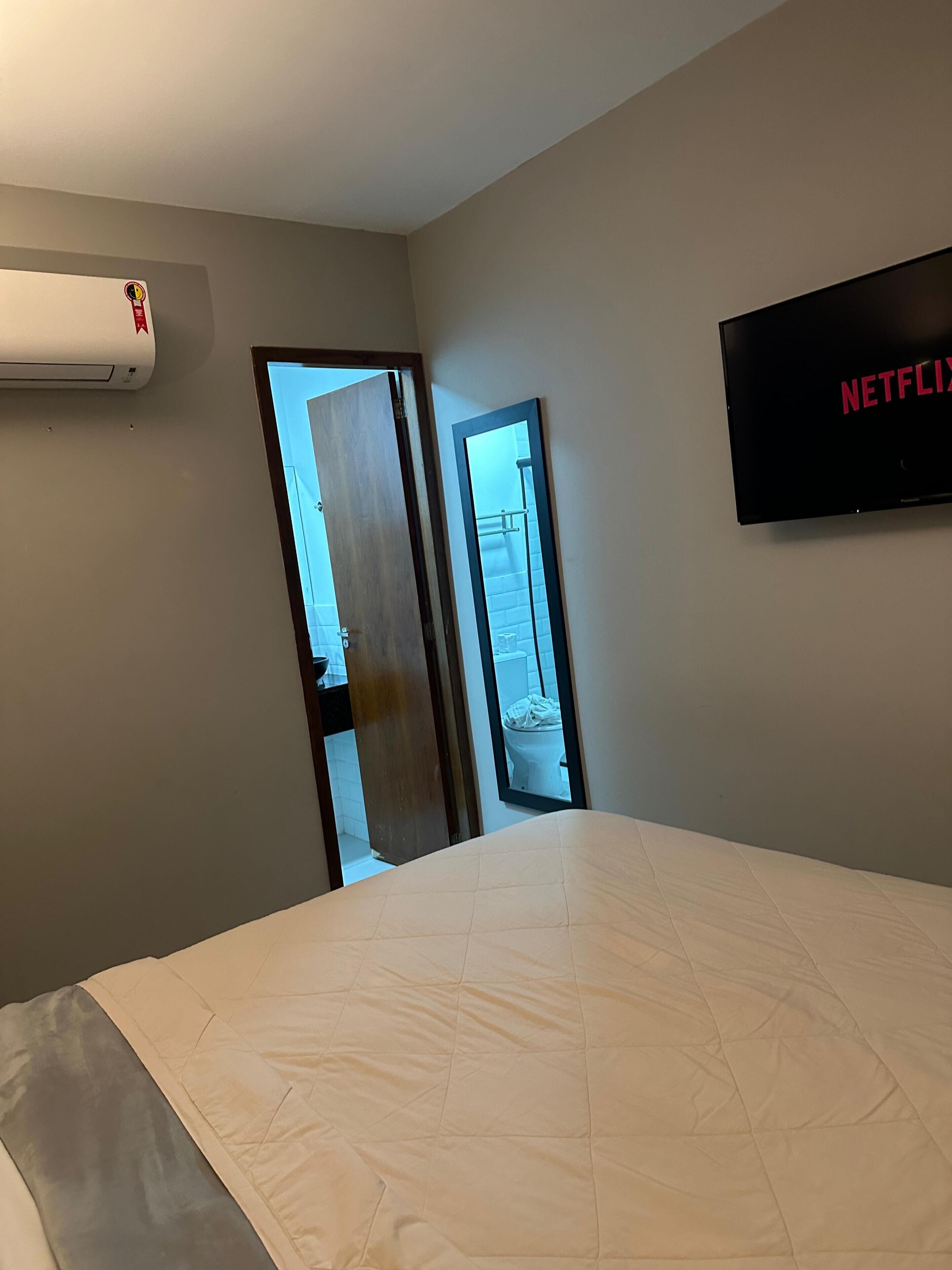 Quarto de hotel com uma cama, ar condicionado, uma televisão exibindo 'NETFLIX' e uma porta que dá para um banheiro.