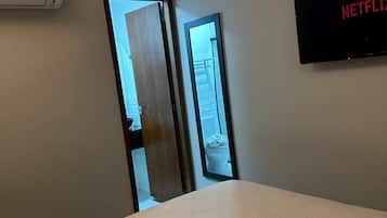 Quarto de hotel com uma cama, ar condicionado, uma televisão exibindo 'NETFLIX' e uma porta que dá para um banheiro.