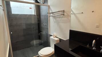 Casa de banho moderna com cabine de chuveiro em vidro, sanita, lavatório preto com espelho e soalho em madeira escura.