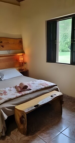 Quarto com cabeceira de madeira, cama com colcha florida, um banco ao pé da cama e janela com vista para o verde.