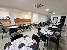 Área de refeições de hotel com mesas preparadas para hóspedes, área de cozinha ao fundo e um extintor de incêndio na parede.