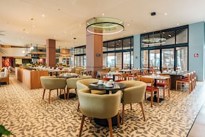 Ampla sala de jantar de hotel, com várias mesas e cadeiras, uma coluna central e janelas grandes que oferecem vistas.