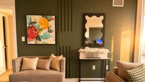 Uma sala de estar com um sofá, uma mesinha de apoio com a placa 'HOME' e um quadro de flores, além de um espelho.