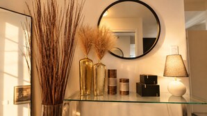 Casa de banho moderna com toucador de vidro, espelho redondo e objetos decorativos, incluindo ervas altas e recipientes cilíndricos.