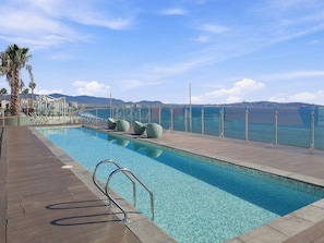 Piscina com vista para o mar e montanhas, rodeada por palmeiras e mobiliário moderno.