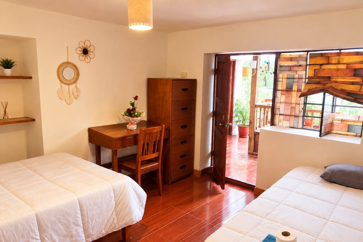 Quarto com duas camas, uma escrivaninha com cadeira, guarda-roupa e janela com vista exterior.