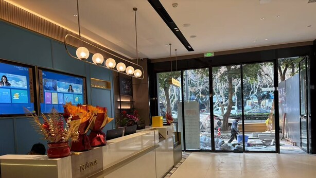 Uma área de recepção de hotel com design moderno, com balcão de atendimento, telas digitais e uma grande porta de vidro que dá para o exterior.