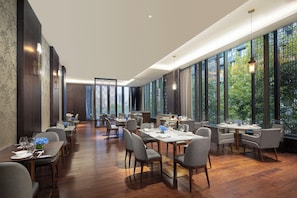 Uma sala de jantar moderna com várias mesas e cadeiras, grandes janelas com vista para o verde e um teto alto com iluminação embutida.